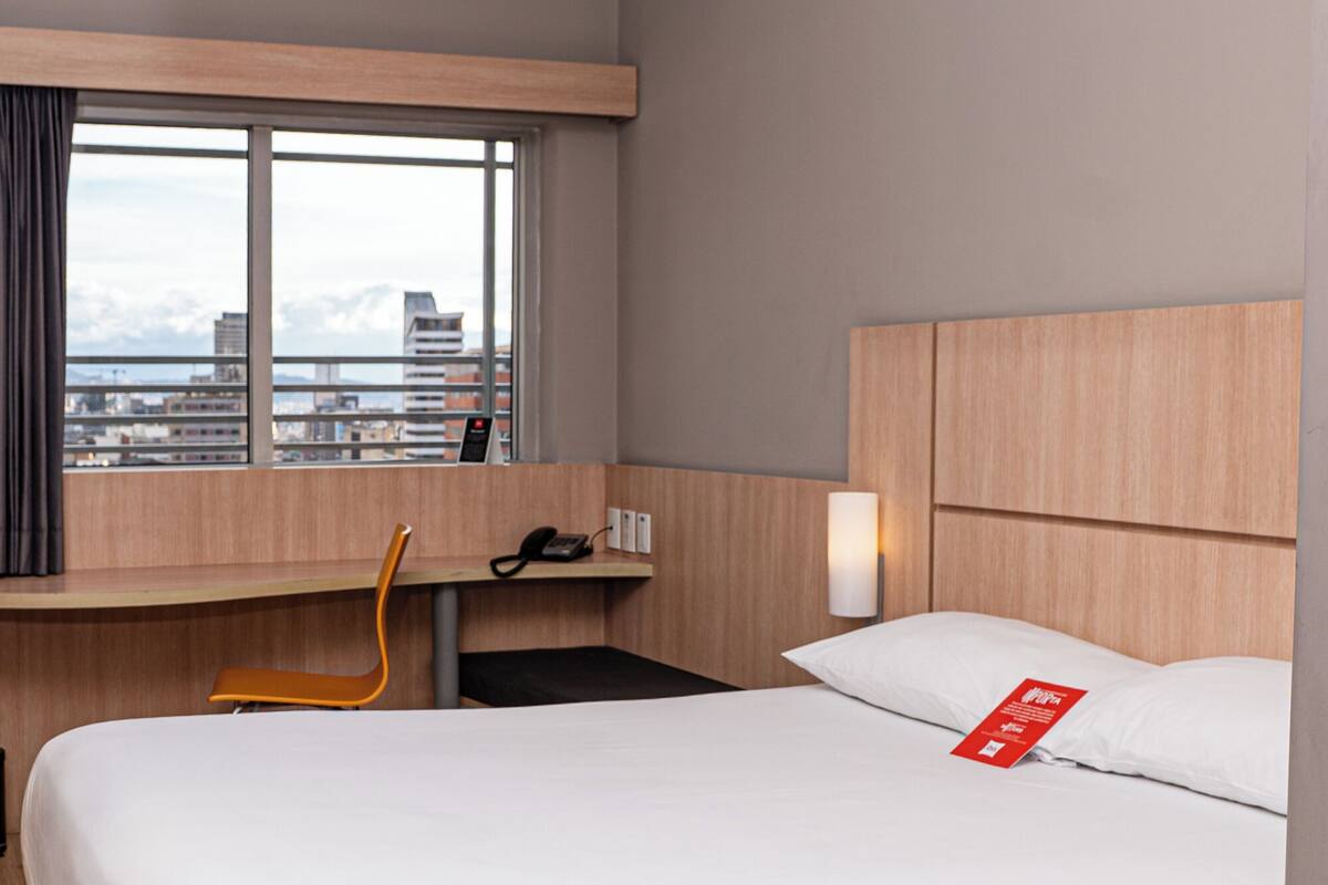 Quarto de hotel com uma cama grande, uma escrivaninha, uma cadeira, uma janela e vista para a cidade, além de uma cabeceira.