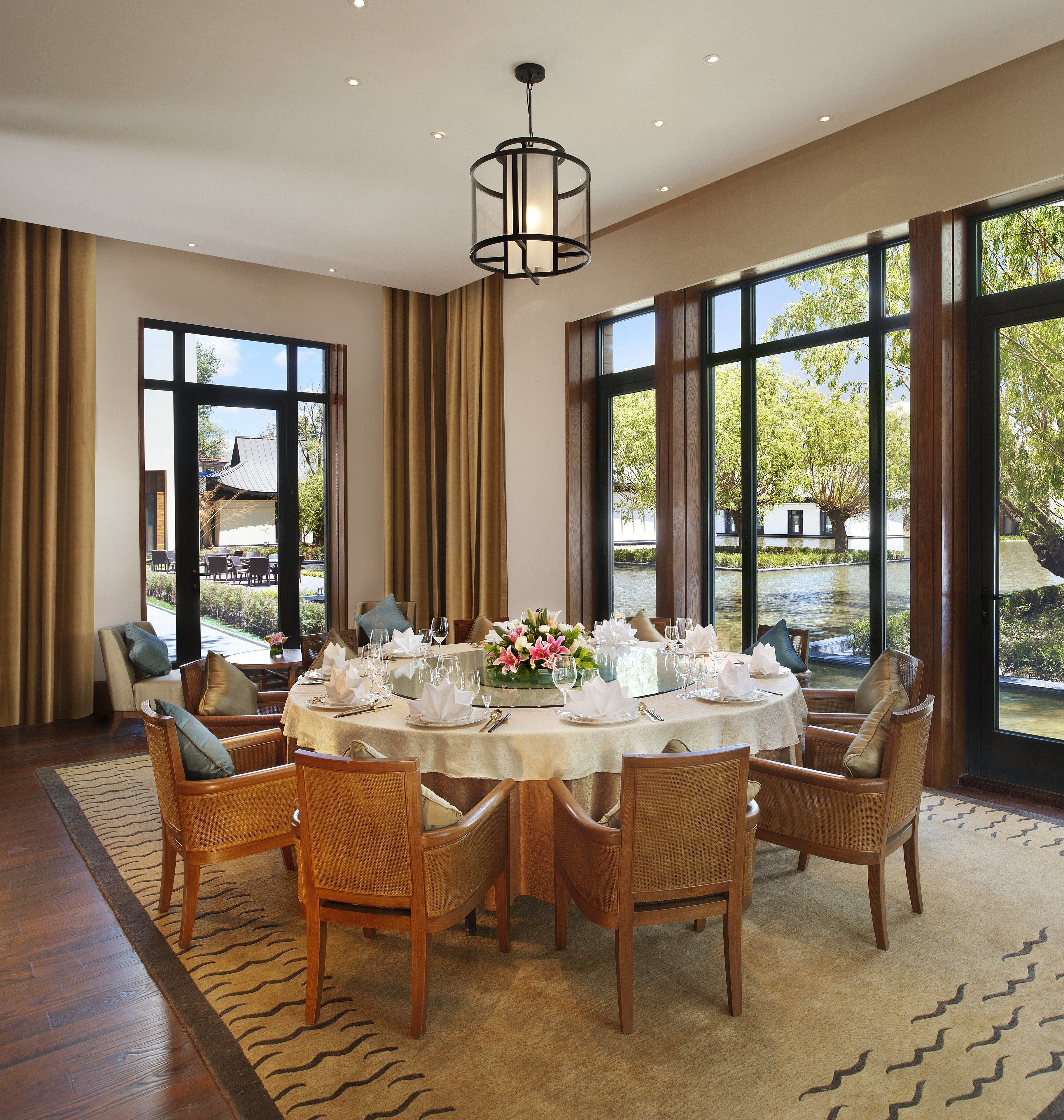 Uma área de refeições com uma mesa redonda posta para seis pessoas, rodeada por cadeiras de vime, grandes janelas com vista para o exterior e um lustre moderno.