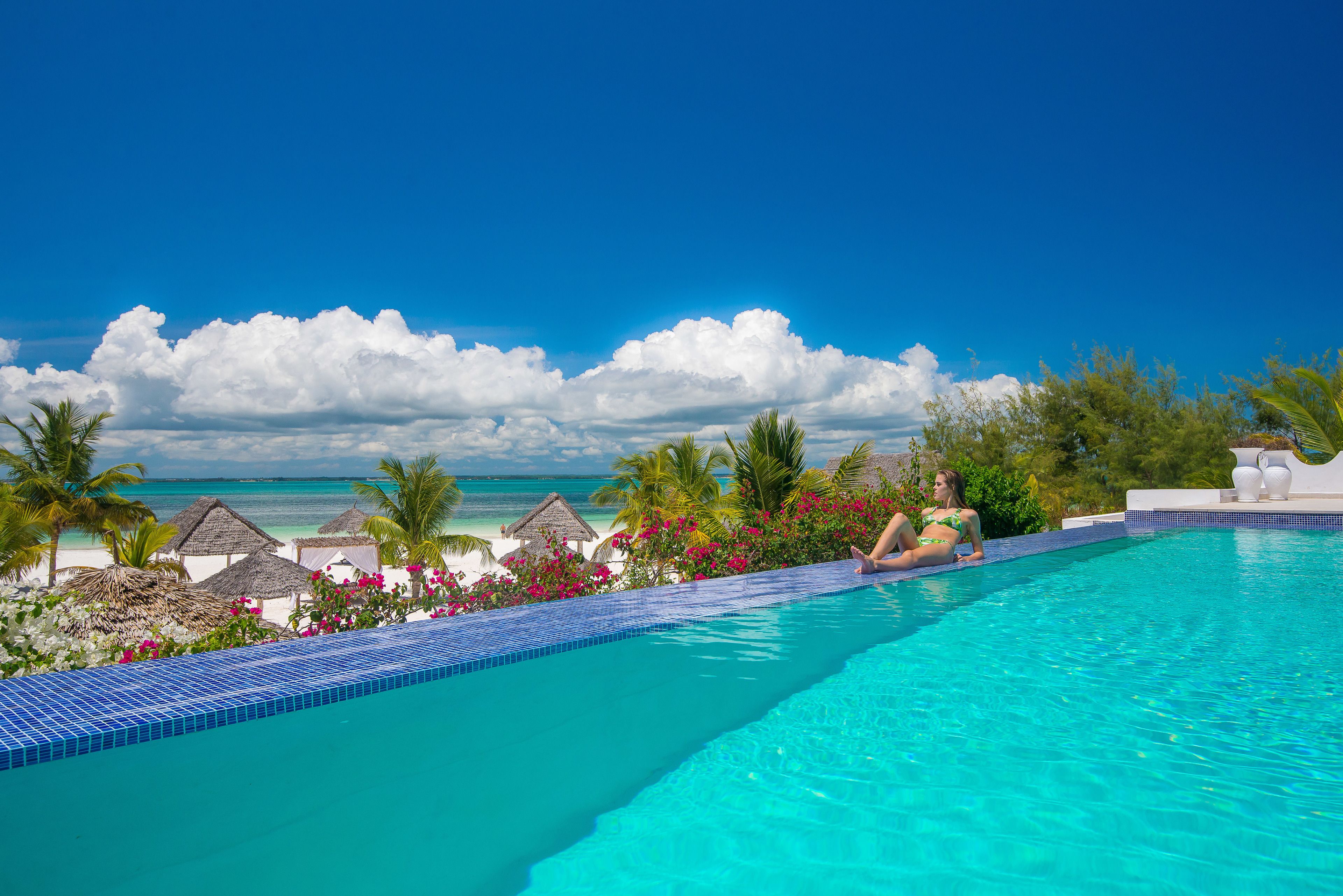 Céu azul límpido, piscina, praia com palmeiras e palafitas.
