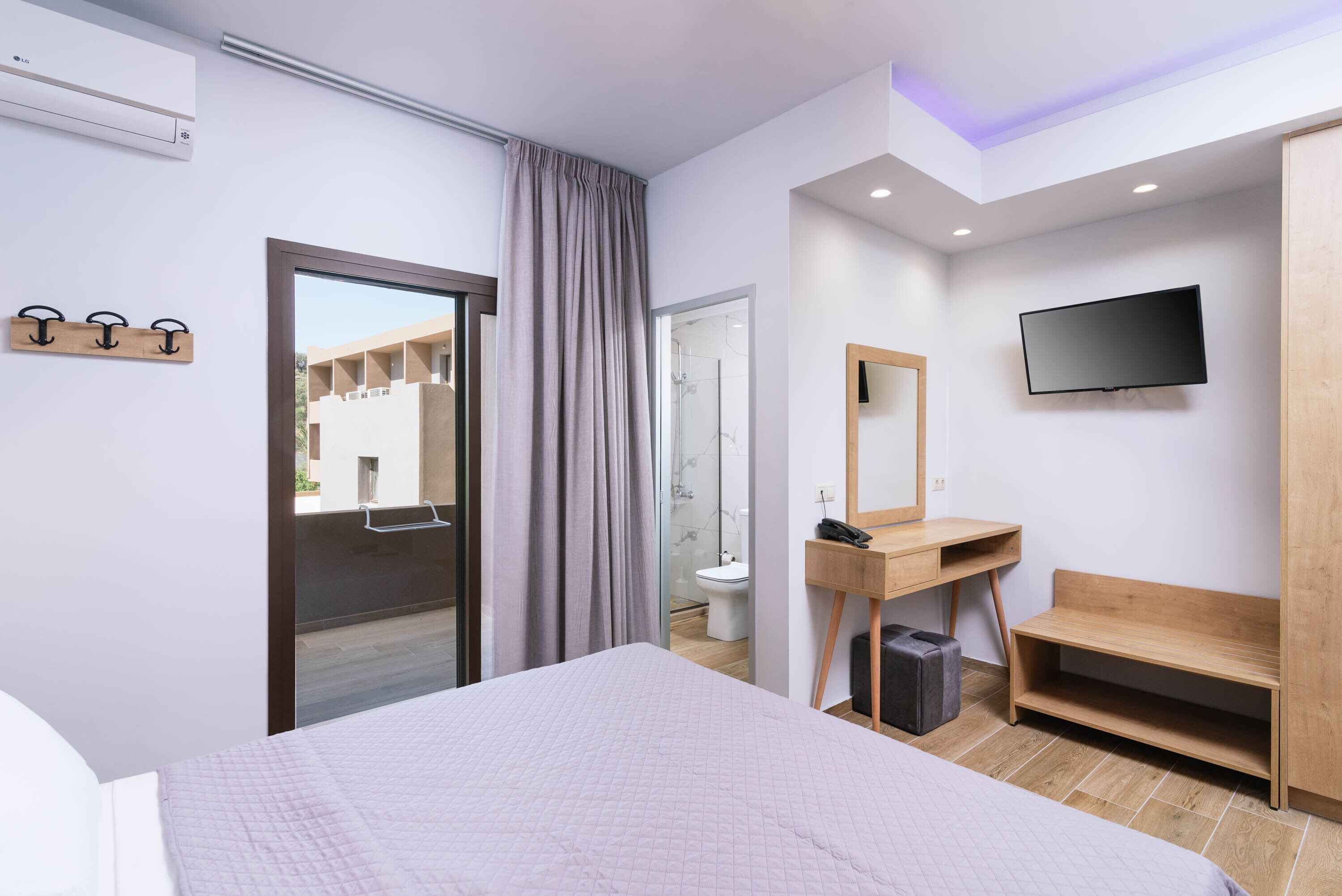 Quarto de hotel moderno com uma cama grande, um banco de madeira, uma mesa com espelho, uma TV fixada na parede e um banheiro visível através de uma porta de vidro.