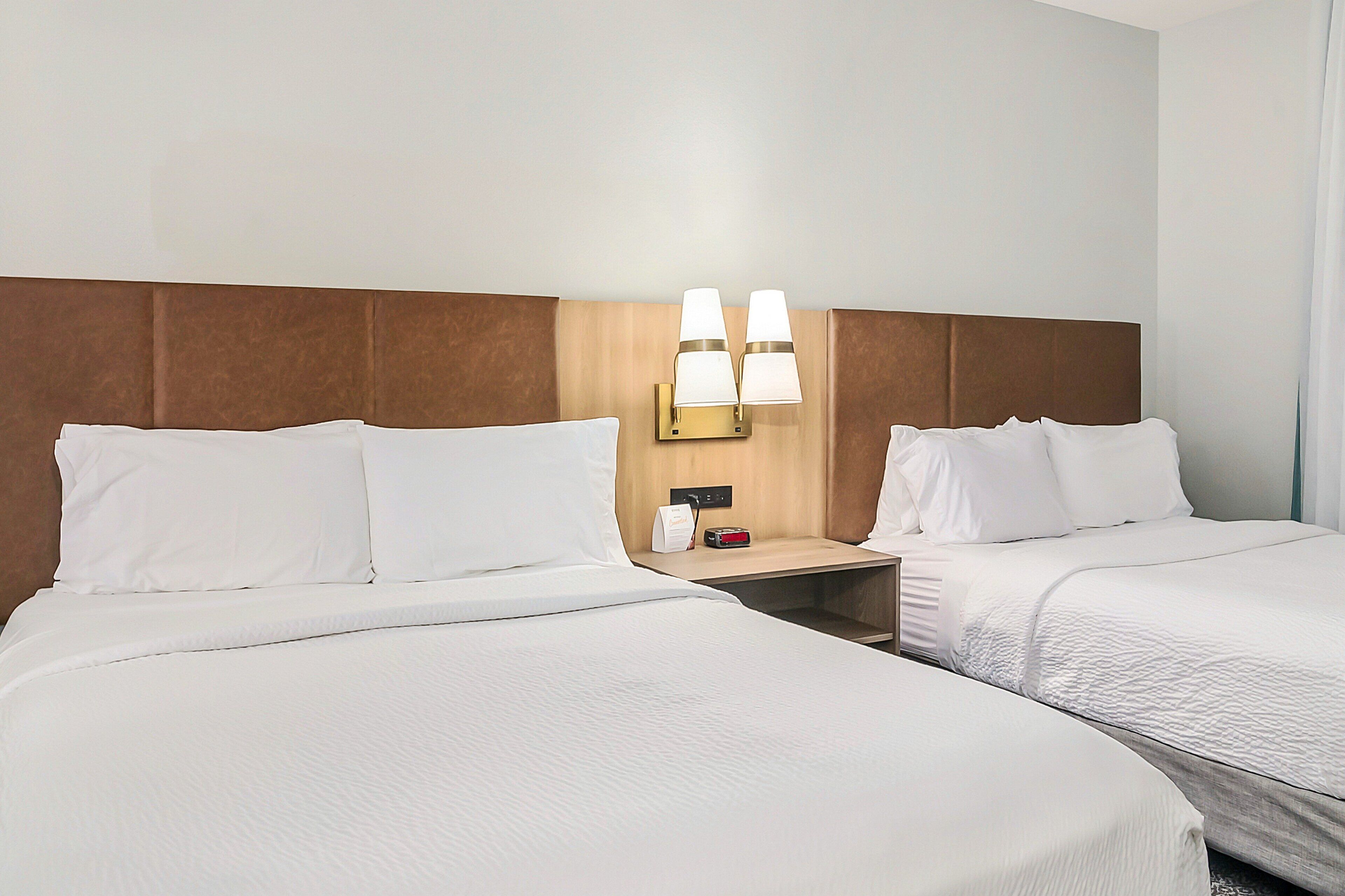 Quarto de hotel com duas camas, uma mesa de cabeceira com luminária e um ponto de luz fixado na parede.