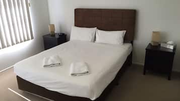 Cama bem arrumada com duas toalhas, cabeceira marrom, almofadas brancas e duas mesas de cabeceira com abajures e caixa de lenços.