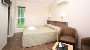 Quarto de hotel bem arrumado com cama, uma cadeira, uma mesinha com um controle remoto e uma janela com vista para o verde.