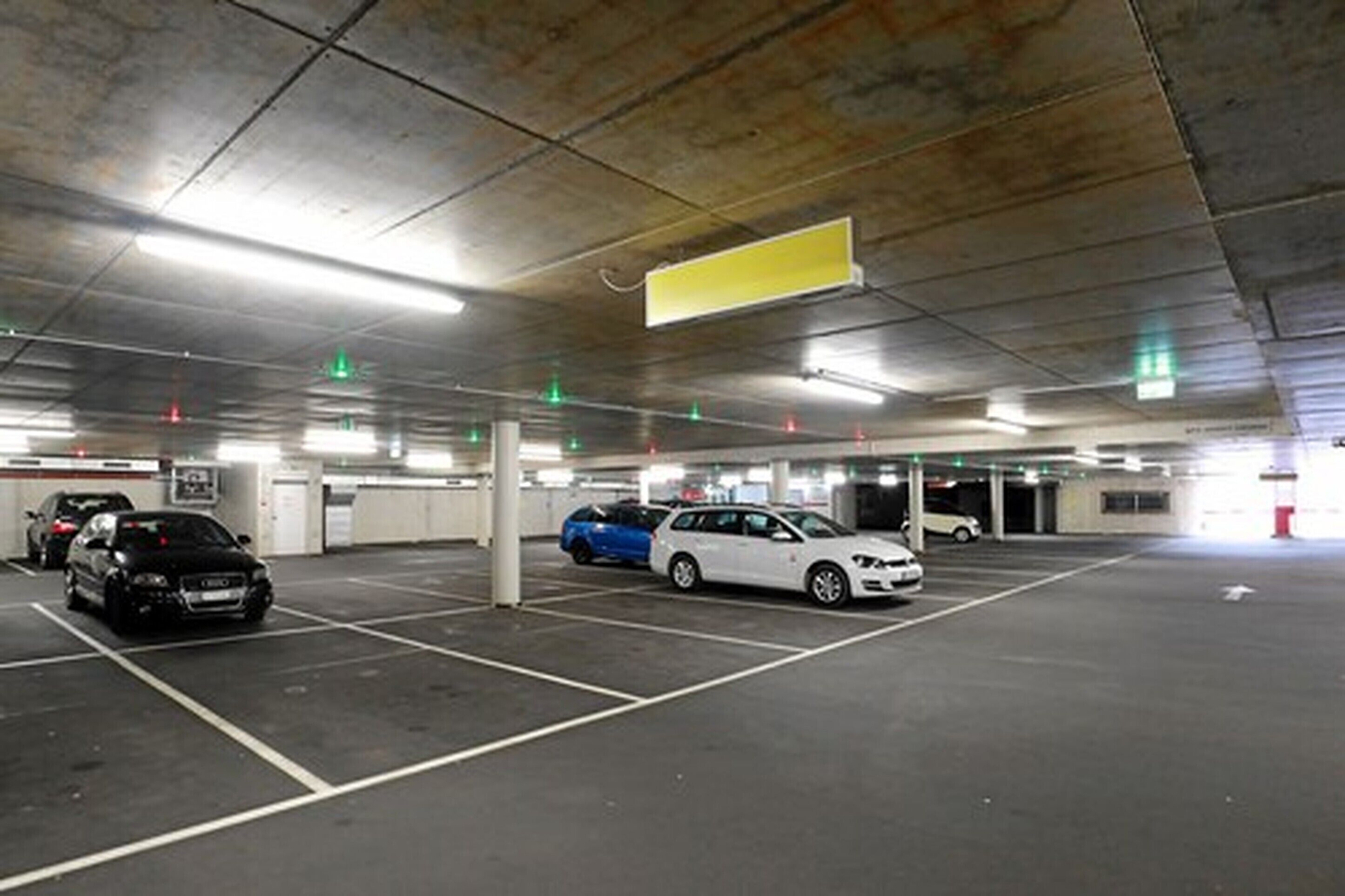 Garagem subterrânea de vários níveis com vários carros estacionados, pilares de concreto e iluminação fluorescente no teto.