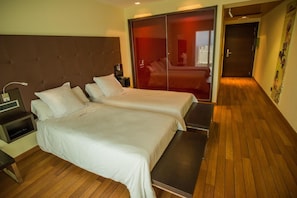 Quarto de hotel com duas camas, piso de madeira, armário com porta deslizante e uma pequena mesa de cabeceira com abajur.