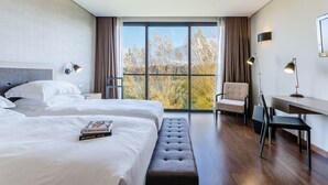 Quarto de hotel com duas camas, uma mesa, uma cadeira e uma janela grande com vista para árvores.