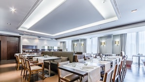 Uma sala de jantar moderna com várias mesas e cadeiras, uma cozinha ao fundo e grandes janelas que oferecem luz natural.