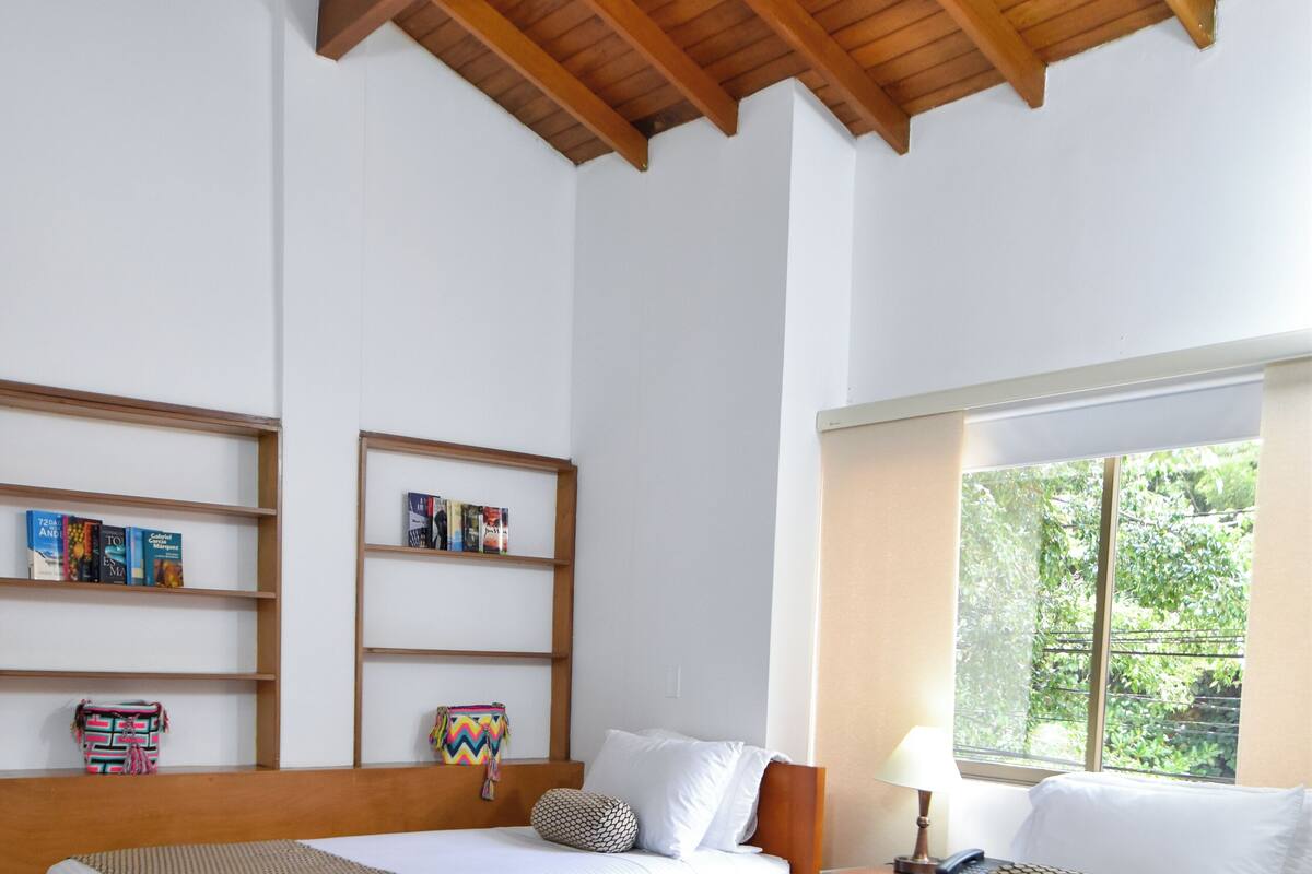 Quarto com duas camas de madeira, uma estante, uma janela com vista para o verde e um abajur em uma mesa de cabeceira.
