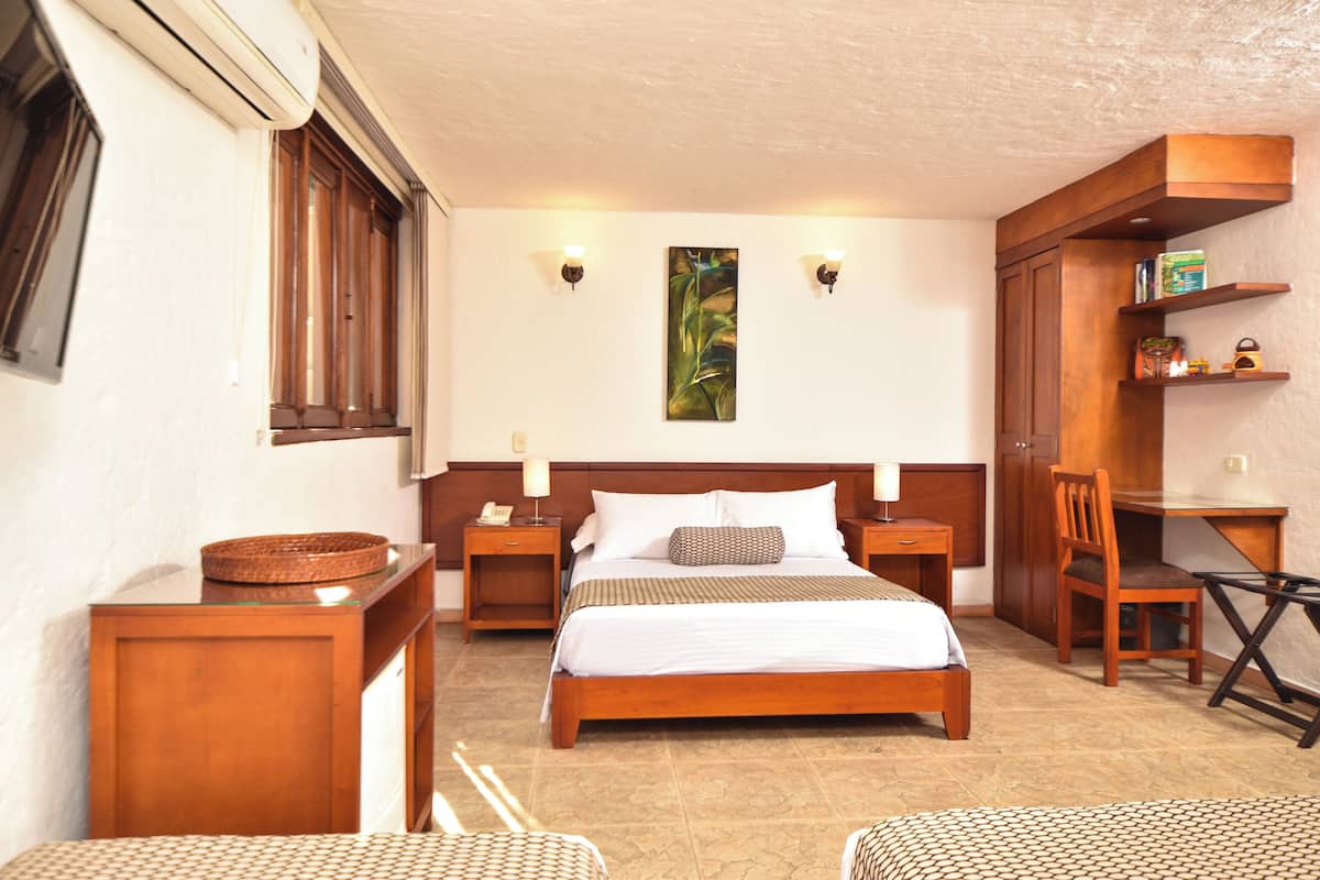 Quarto de hotel com uma cama grande, cabeceira de madeira, duas mesas de cabeceira, uma escrivaninha, uma cadeira e uma estante com livros e um rádio.