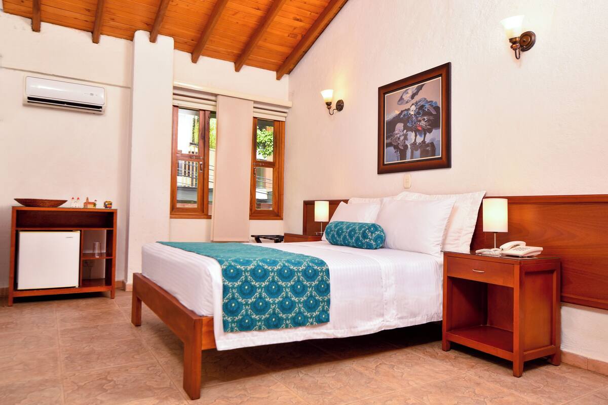 Um quarto com cama de madeira, mesa de cabeceira com abajur, um pequeno criado-mudo de madeira, ar condicionado fixado na parede e uma porta que dá para área externa.