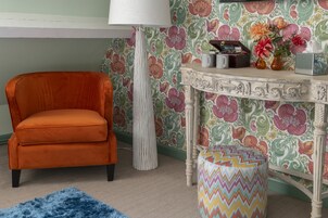 Quarto com poltrona laranja acolchoada, luminária de chão branca alta, mesinha decorativa com um vaso de flores e um banquinho estampado.