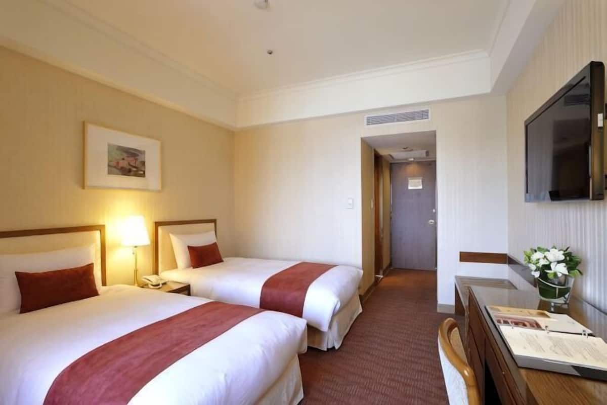 Quarto de hotel com duas camas, uma escrivaninha, uma cadeira, uma televisão e uma porta que dá para outro cômodo.