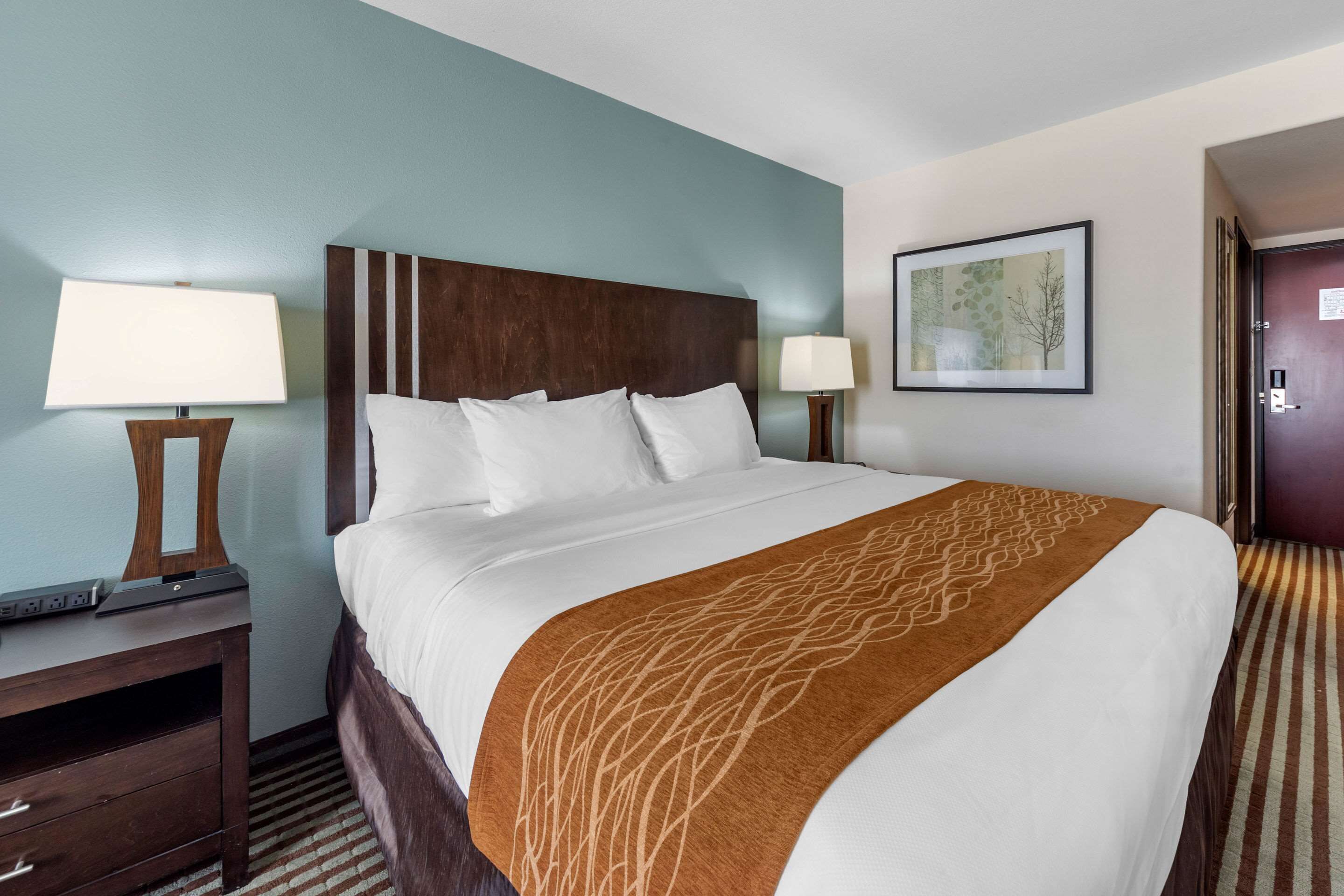 Quarto de hotel com uma cama grande, duas luminárias de cabeceira, uma mesinha de cabeceira e uma moldura com um quadro na parede.
