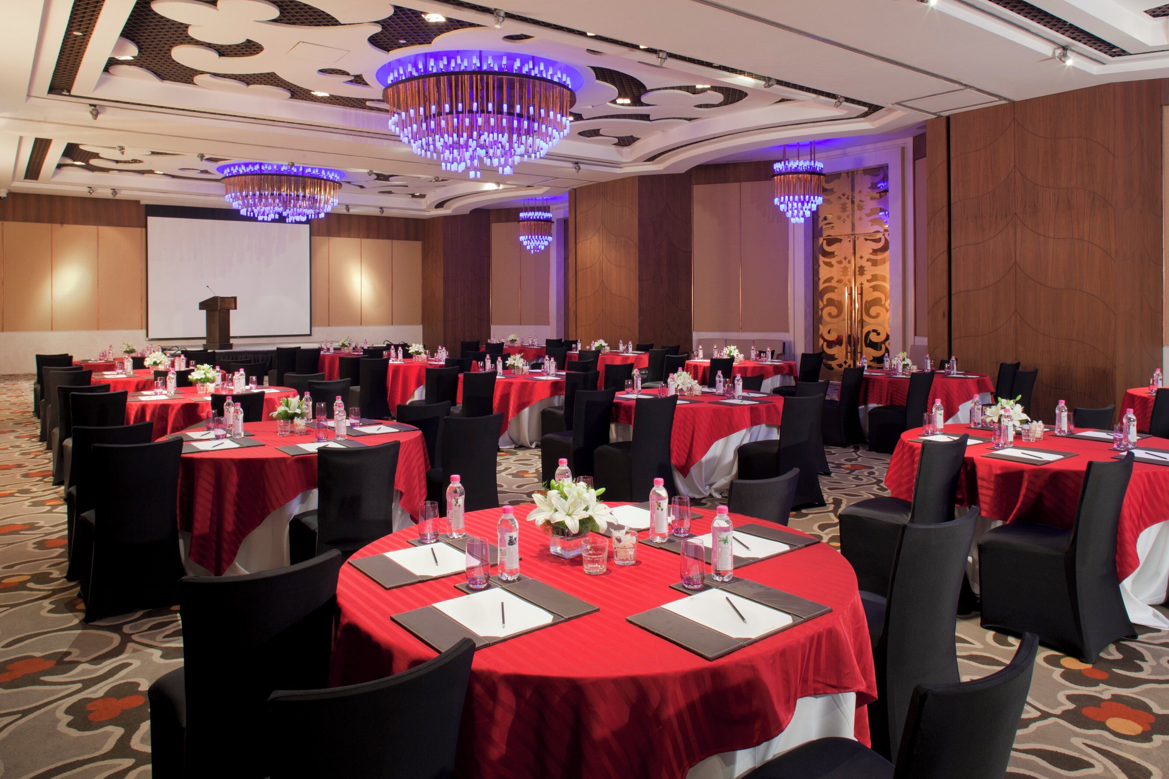 Sala de conferências com mesas redondas, cadeiras pretas e toalhas de mesa vermelhas.