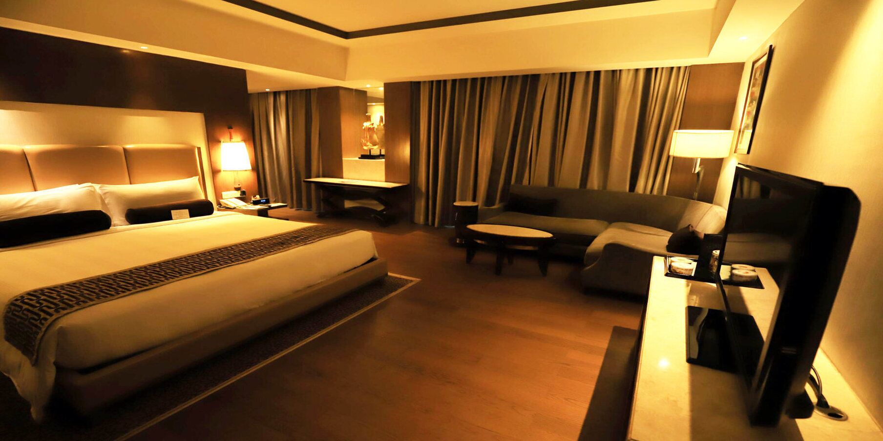 Quarto de hotel com uma cama grande, um sofá, uma televisão e uma mesa de trabalho com luminária.