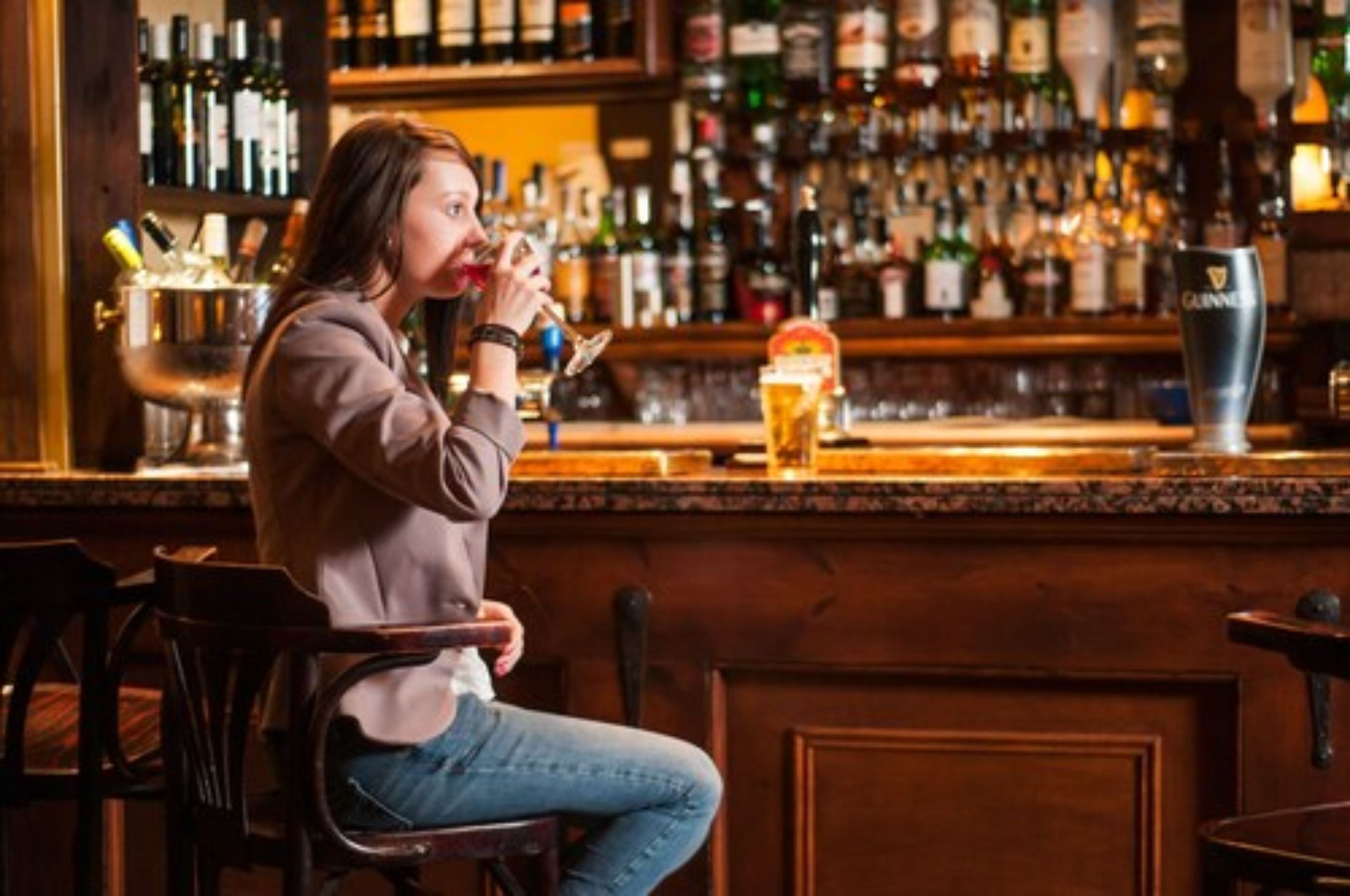 Uma pessoa sentada em um bar, com diversas garrafas e uma torneira ao fundo.