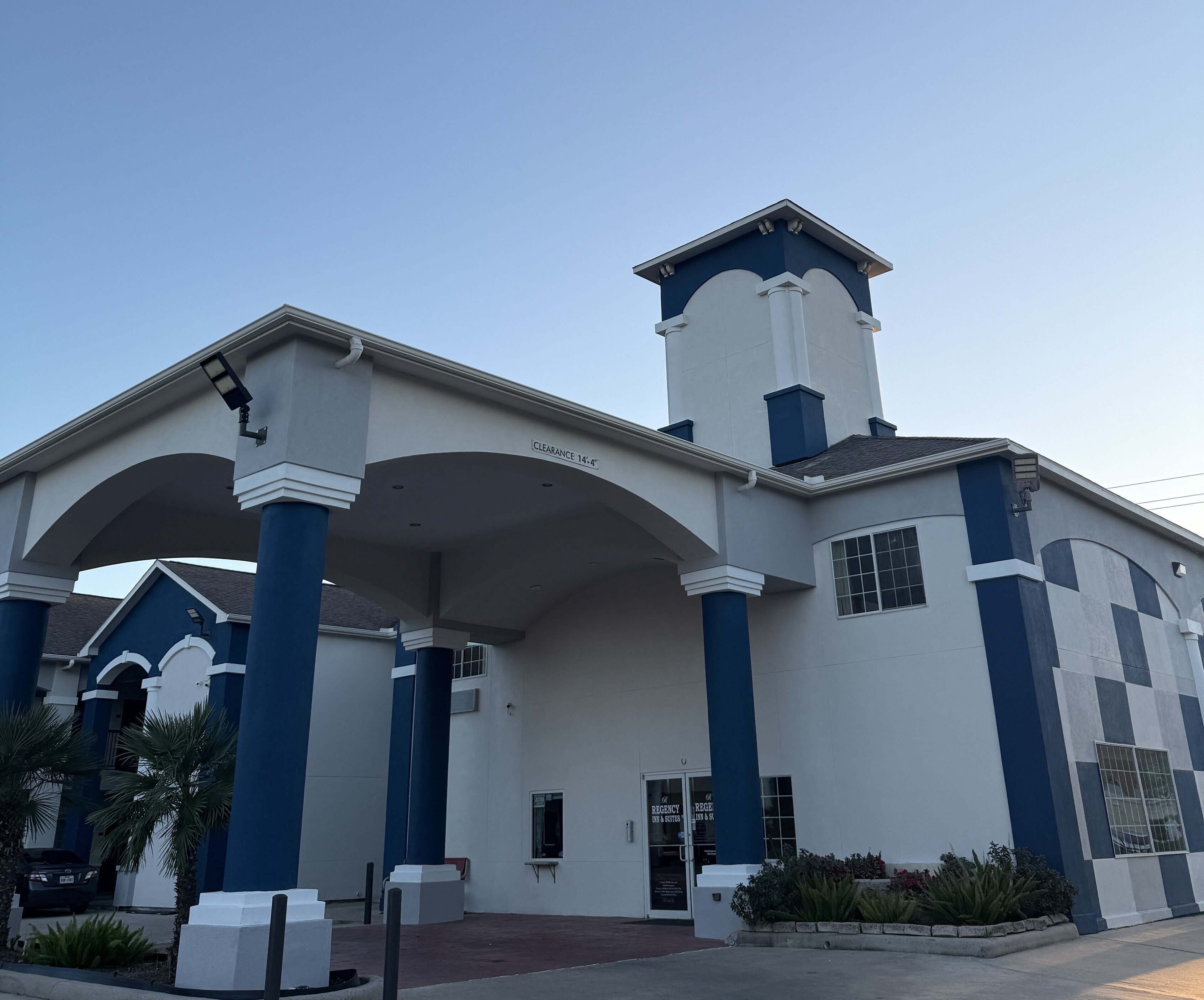 Um edifício com fachada branca e azul, uma entrada central com marquise e uma pequena estrutura semelhante a torre à direita.