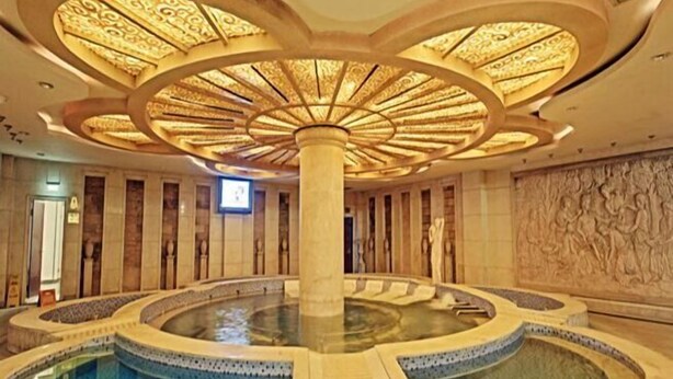 Uma área de piscina interior luxuosa, com um pilar central, teto ornamentado e paredes de pedra.