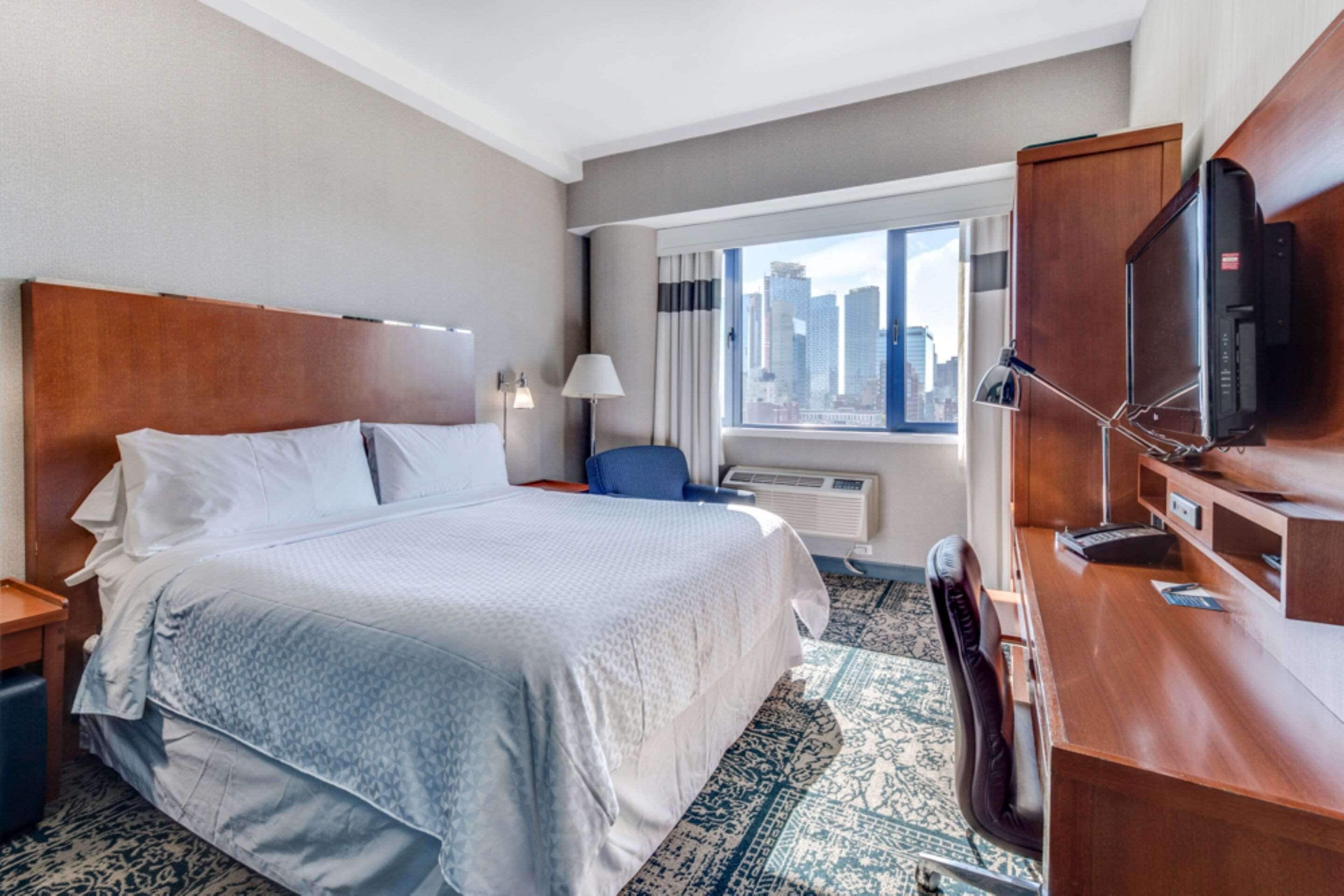 Quarto de hotel com uma cama grande, uma escrivaninha com computador, televisão e janela com vista para a cidade.
