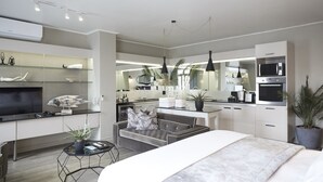 Cozinha moderna e bem iluminada com ilha central, área de jantar com mesa de vidro e uma cama grande com cabeceira branca.
