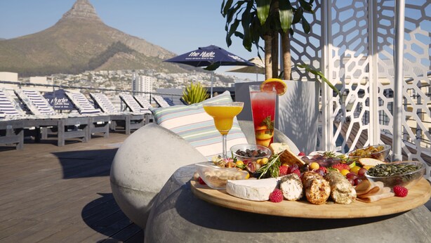Uma mesa de jantar com diversos alimentos e bebidas, vista para a praia e cadeiras de descanso ao fundo.