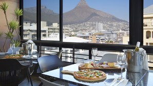 Uma mesa de jantar com pizza, copos de vinho e uma garrafa de champanhe, com vista para uma paisagem urbana e uma montanha.