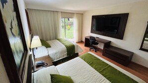 Quarto de hotel com duas camas, uma escrivaninha, uma cadeira, uma televisão e uma janela com cortinas.