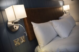 Quarto de hotel com cama, almofadas brancas, cabeceira de madeira e luminária fixada na parede.