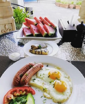 Uma mesa de café da manhã com pratos de salsichas, ovos e legumes, e uma bandeja de melancia em fatias.