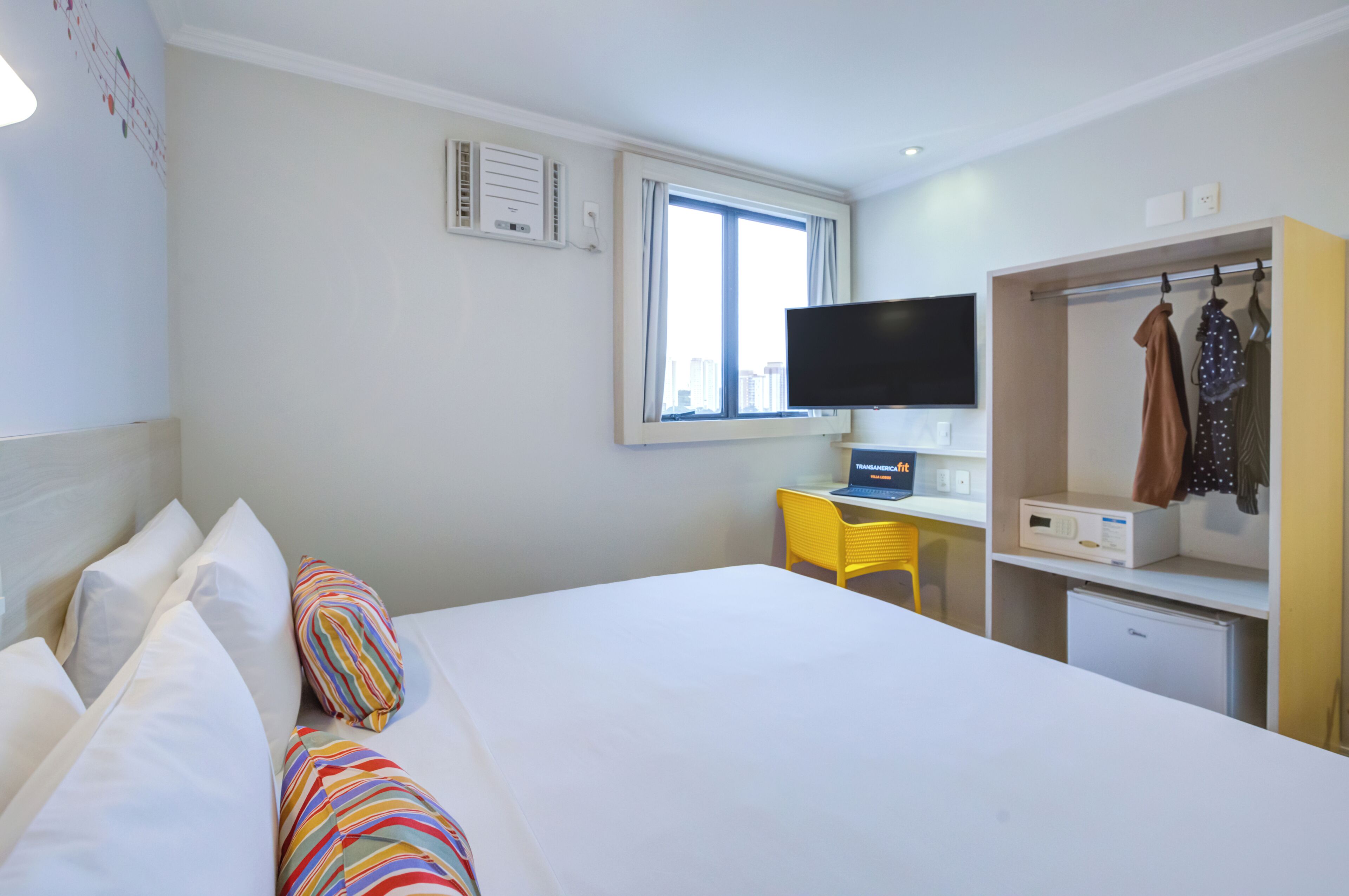 Quarto de hotel com cama, secretária, computador, televisão e armário.
