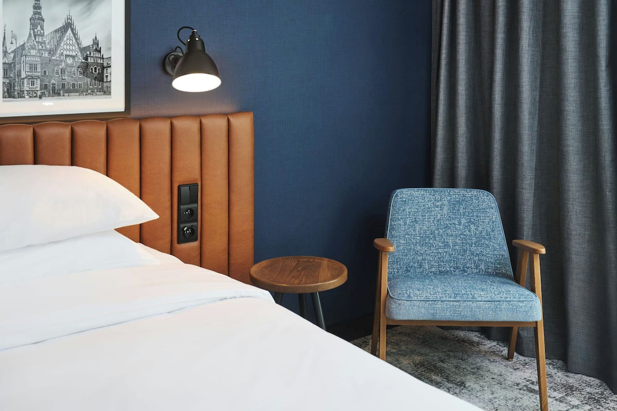 Quarto de hotel com uma cama, uma luminária fixada na parede, uma mesa redonda pequena, uma poltrona azul estofada e um quadro na parede.