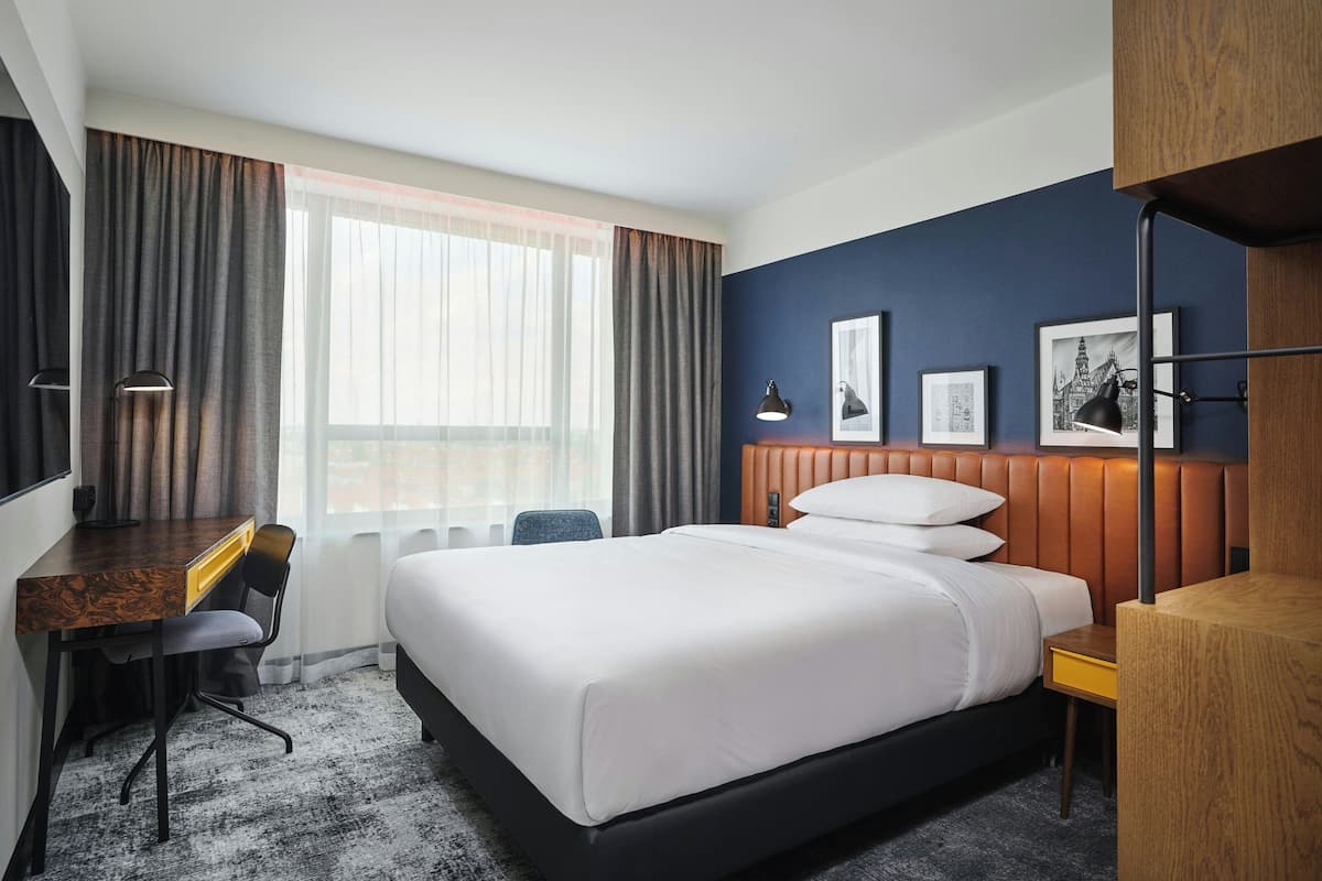 Quarto de hotel com cama, escrivaninha, cadeira e uma janela grande com cortinas.