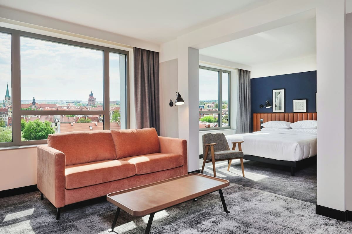 Quarto de hotel com uma janela grande, um sofá, uma cama, uma cadeira e uma mesinha.