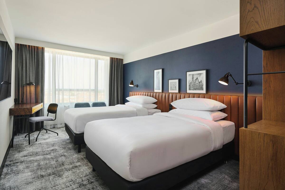 Quarto de hotel com duas camas, uma escrivaninha, uma cadeira e uma janela grande.
