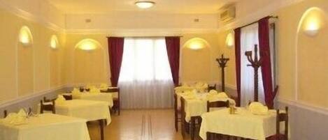 Uma sala de jantar de hotel com várias mesas preparadas para refeições, um candelabro e uma grande janela com cortinas.