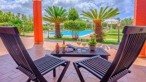 Um pátio com duas cadeiras pretas, uma mesa com garrafas e copos, uma piscina e palmeiras.