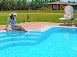 Uma área de piscina com uma pessoa sentada à beira da piscina, uma espreguiçadeira e um edifício ao fundo.