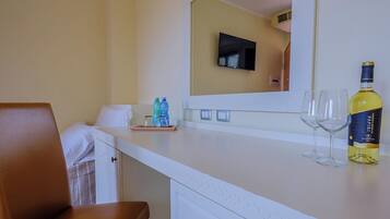 Quarto de hotel com cômoda branca, poltrona de couro marrom, espelho, televisão, duas garrafas de água e dois copos de vinho vazios.