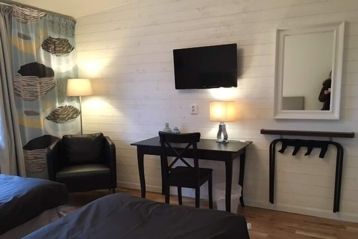 Quarto de hotel com cama, escrivaninha, cadeira, televisão, abajur e espelho.
