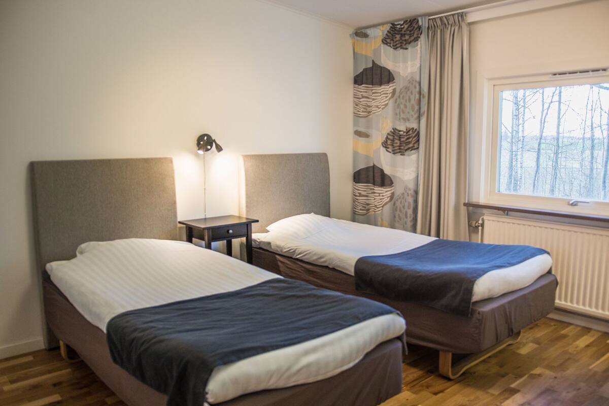 Quarto de hotel com duas camas, uma mesinha de cabeceira, uma lâmpada, uma janela com vista para árvores e um radiador.
