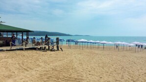 Uma praia com uma estrutura de madeira, possivelmente um bar ou restaurante, e fileiras de guarda-sóis.