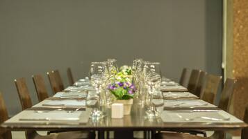 Uma mesa de jantar comprida disposta com cadeiras, um arranjo floral no centro e um objeto branco pequeno sobre a mesa.