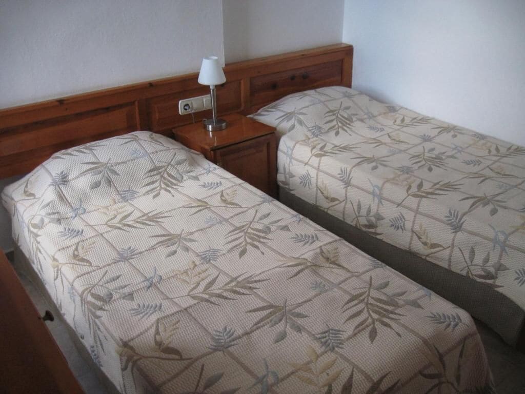 Quarto com duas camas de solteiro, roupa de cama com padrão de folhas, mesa de cabeceira com abajur e cabeceira de madeira.