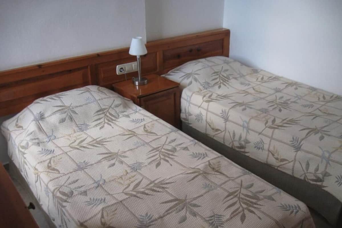 Quarto com duas camas de solteiro, roupa de cama com padrão de folhas, mesa de cabeceira com abajur e cabeceira de madeira.