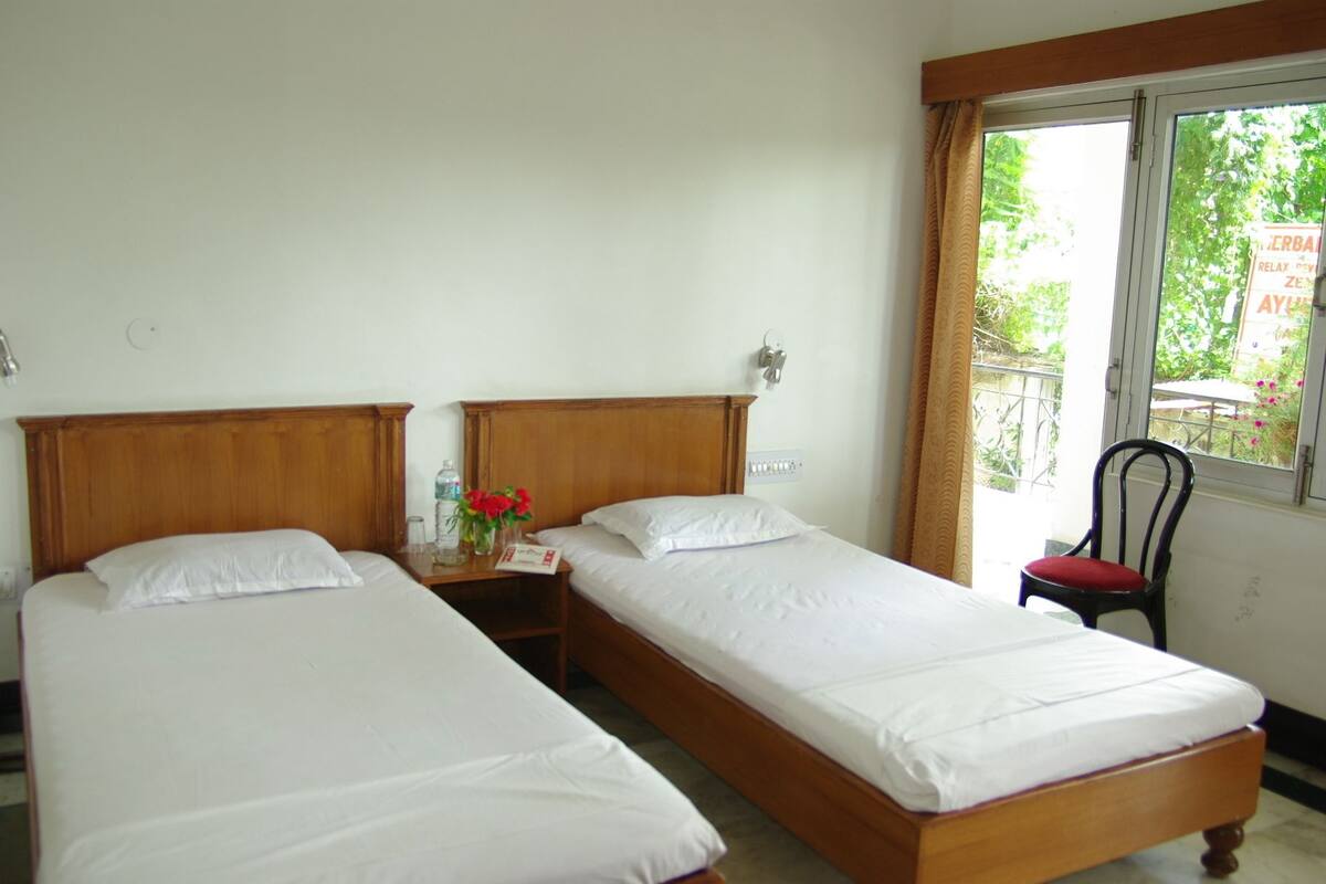 Quarto com duas camas de solteiro, mesa de cabeceira com uma garrafa de água e um vaso de flores, uma cadeira e janela com vista para o verde.