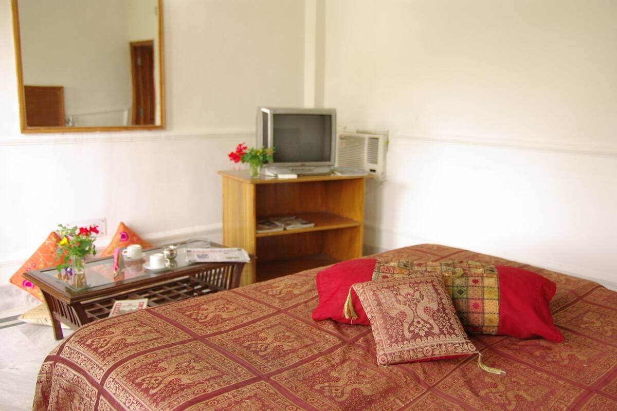 Quarto de hotel com uma cama, televisão sobre um suporte de madeira, uma mesinha com um vaso e um jornal, e um espelho na parede.