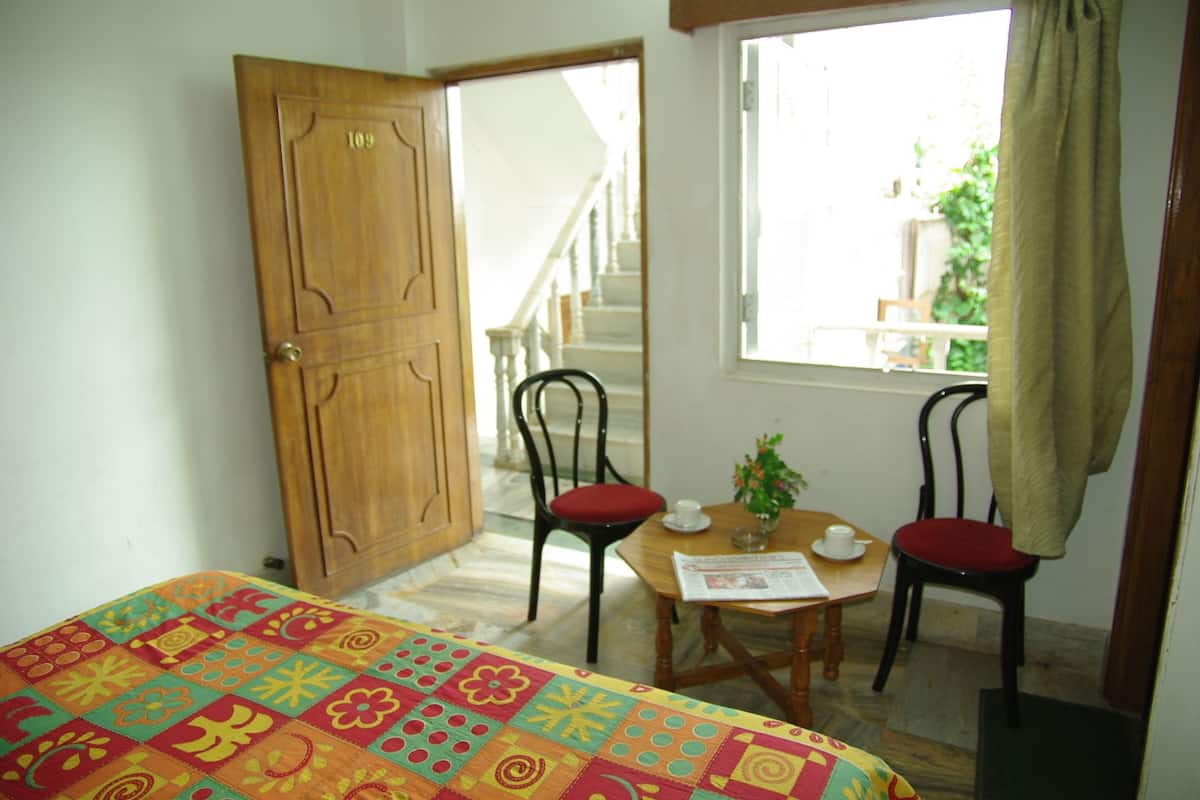 Quarto com porta de madeira, cama com colcha colorida, duas cadeiras, uma mesa pequena com uma planta e janela com vista para uma escadaria e vegetação lá fora.