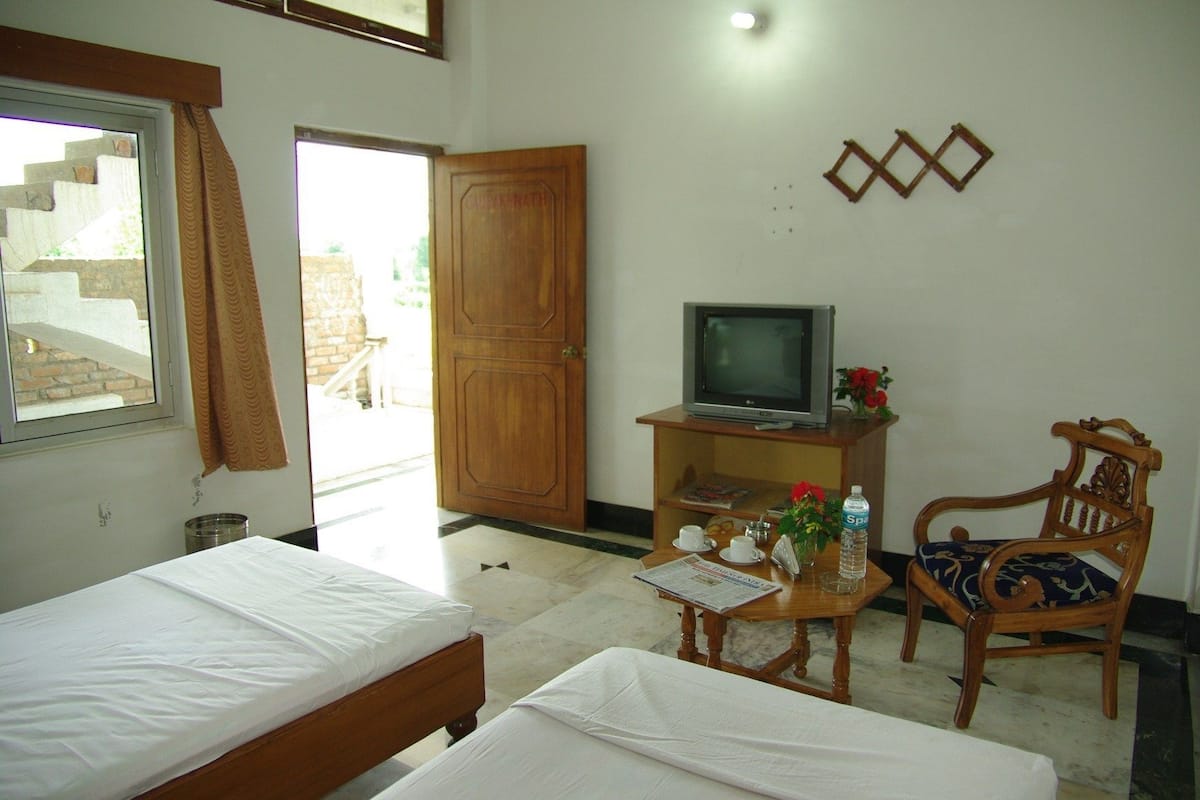 Quarto de hotel com duas camas de solteiro, uma televisão sobre um suporte de madeira, uma cadeira, uma mesa pequena com um jornal e uma janela com cortinas.