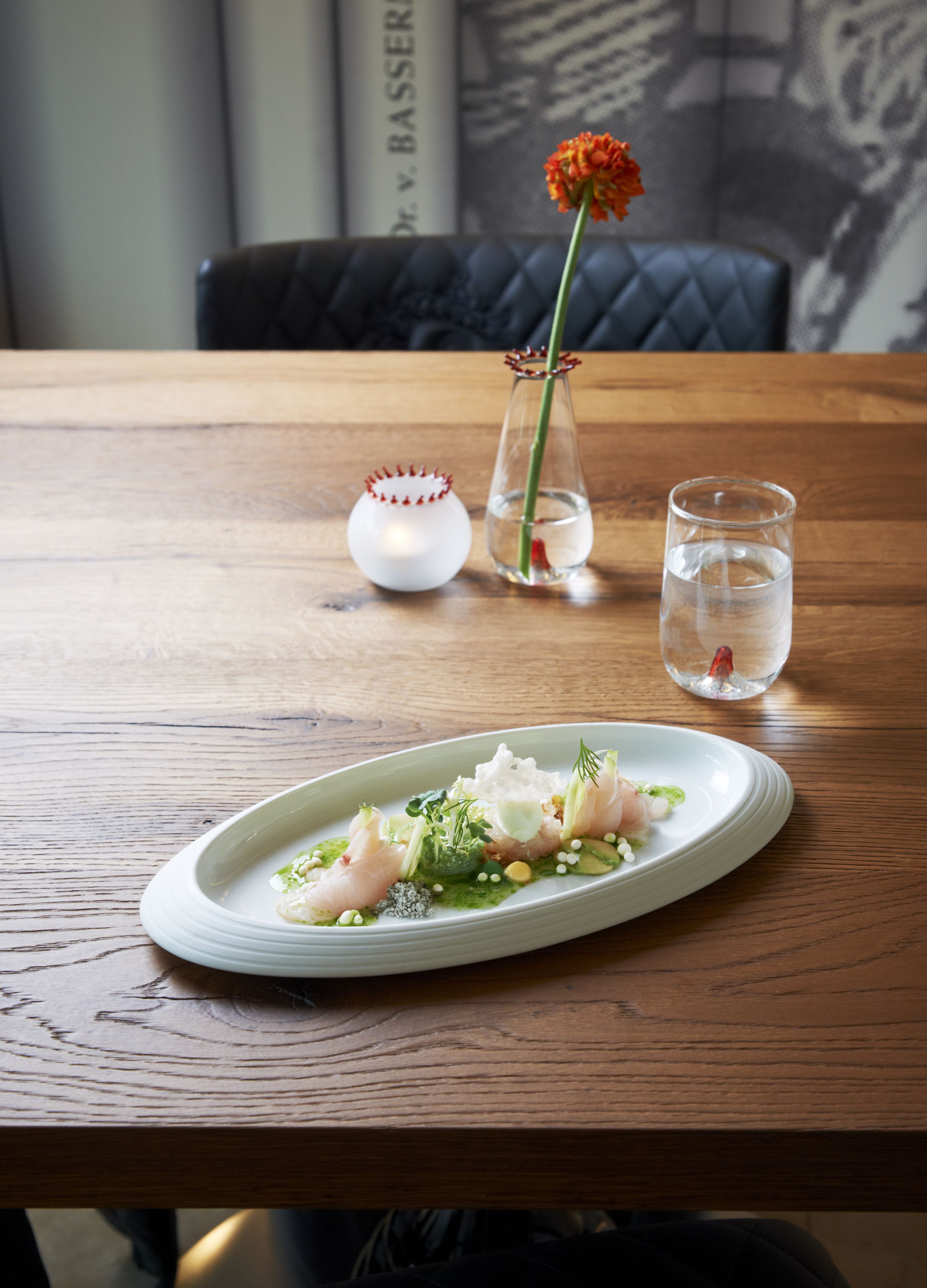 Uma mesa de madeira com um prato de comida, um copo de água, um vaso com uma flor e um objeto decorativo.