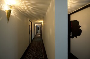 Corredor com carpete estampado, arandelas na parede e uma porta com número.